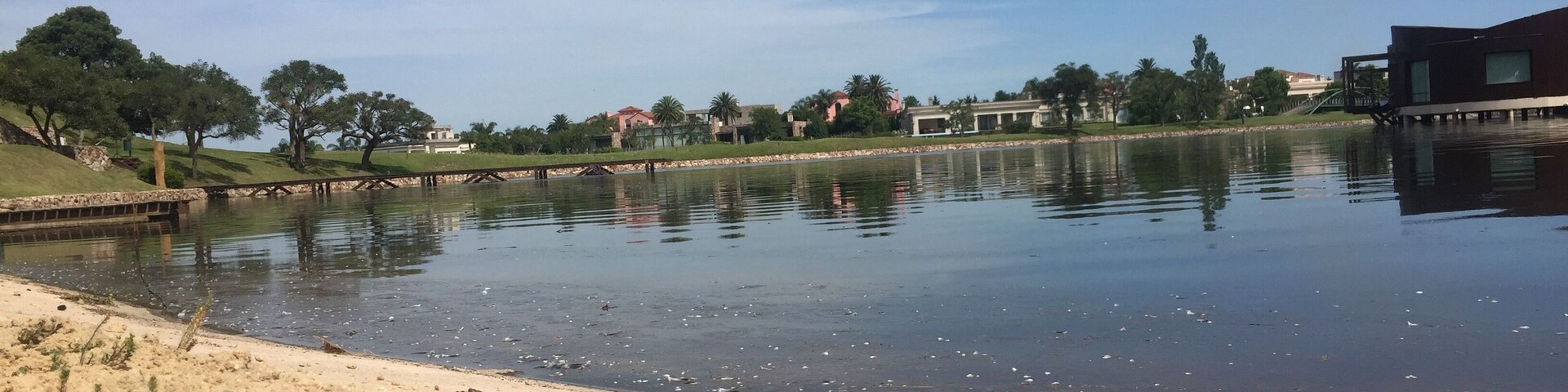 Um lago calmo com margem arenosa, rodeado por árvores e edifícios.