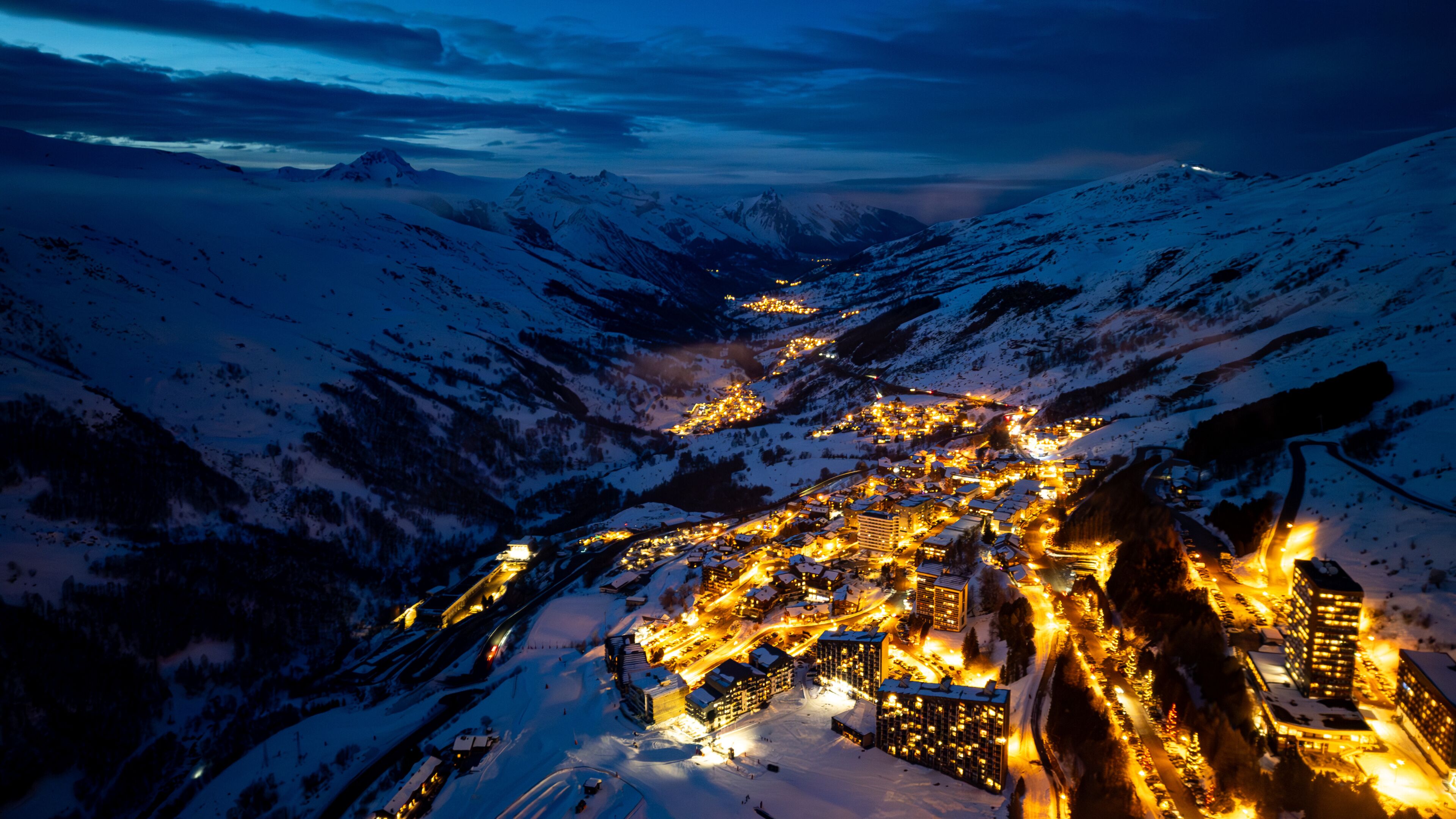 Uma cidade nas montanhas coberta de neve à noite, com edifícios iluminados e estradas sinuosas.
