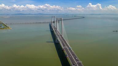 Uma longa ponte suspensa atravessa uma vasta extensão de água, com céu claro e montanhas ao longe.