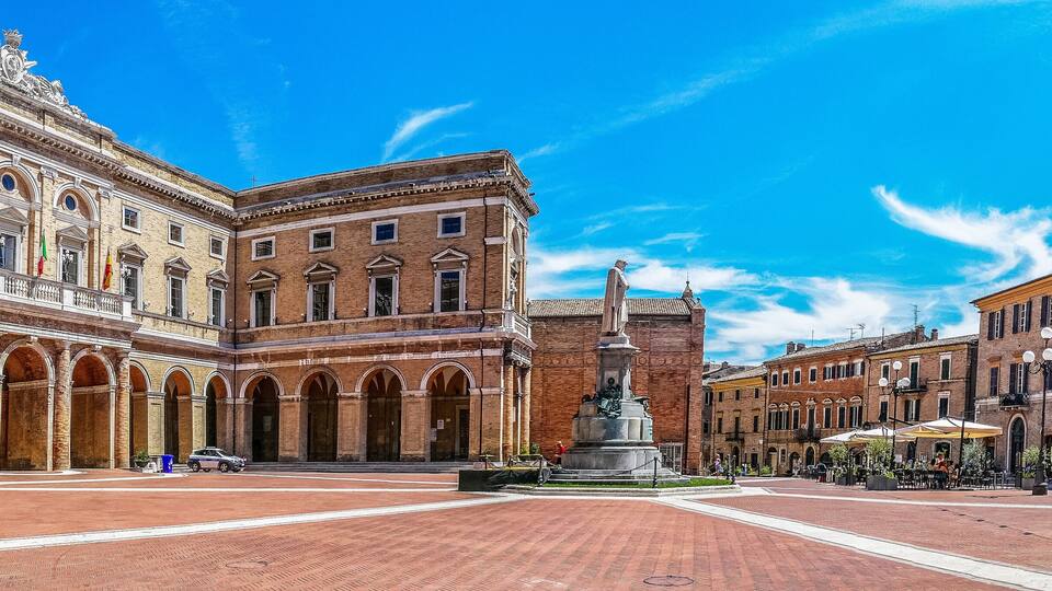 Uma praça histórica com arquitetura clássica, uma estátua central e edifícios ao redor.