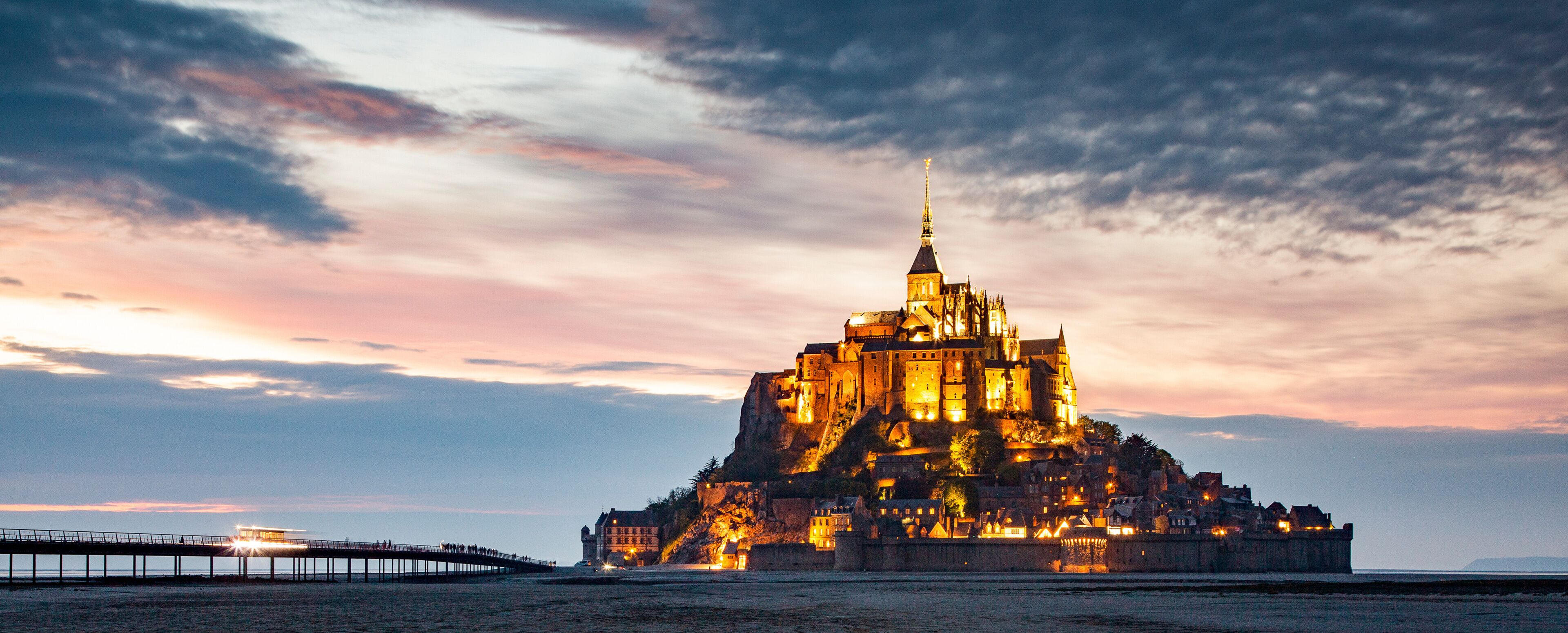 Um castelo histórico numa colina com vista para uma praia, com um cais que se estende para a água.