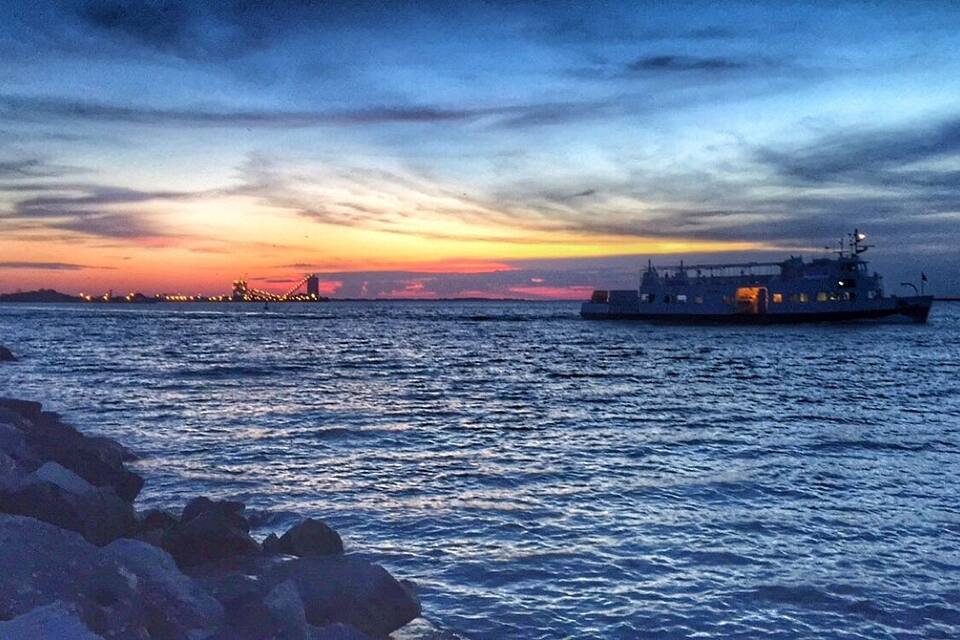 Pôr do sol sobre a água com um grande navio e um litoral rochoso.