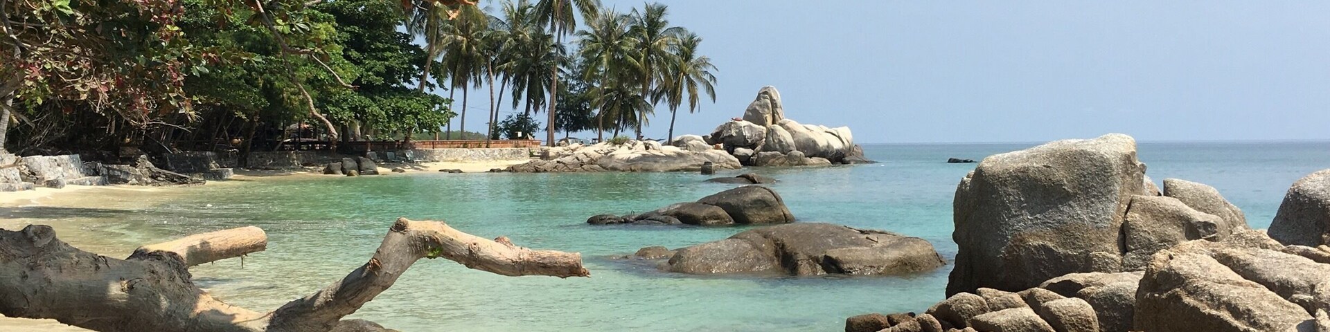Uma praia tropical com pedras grandes, água azul-turquesa cristalina e palmeiras.