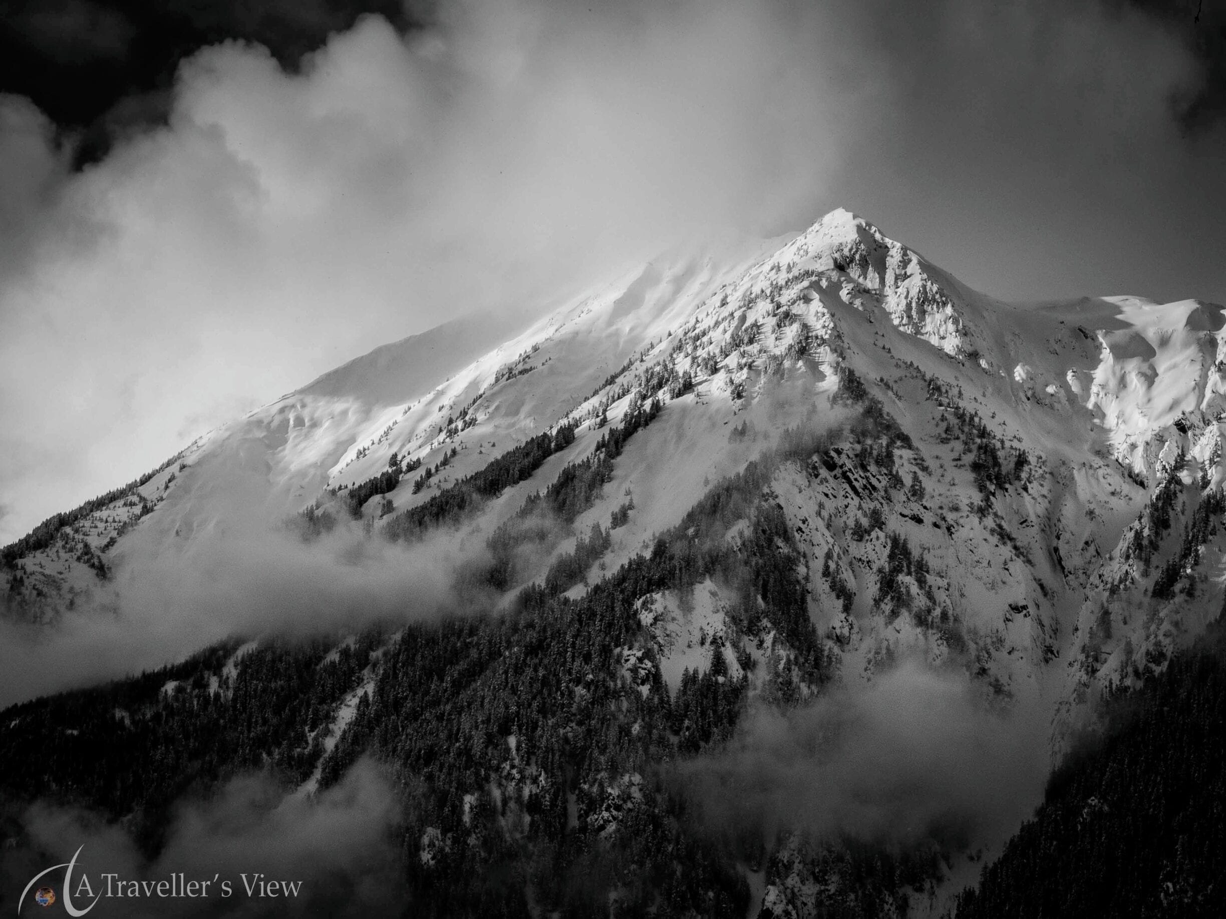 Montanha coberta de neve com pico, encostas arborizadas e nuvens na base.