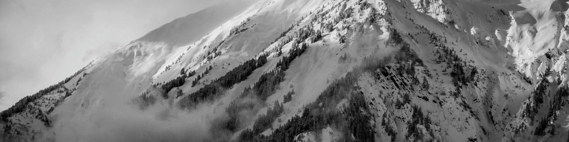 Montanha coberta de neve com pico, encostas arborizadas e nuvens na base.