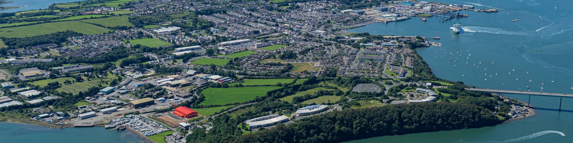 Vista aérea de uma cidade costeira com um porto, uma ponte e campos verdes ao redor.