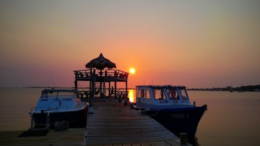 Um pôr do sol sobre águas calmas, com um cais que leva a uma construção com telhado de palha. Dois barcos estão atracados no cais, um deles com a inscrição 'Alton's Dive Center' e o outro 'NAIDA J'.