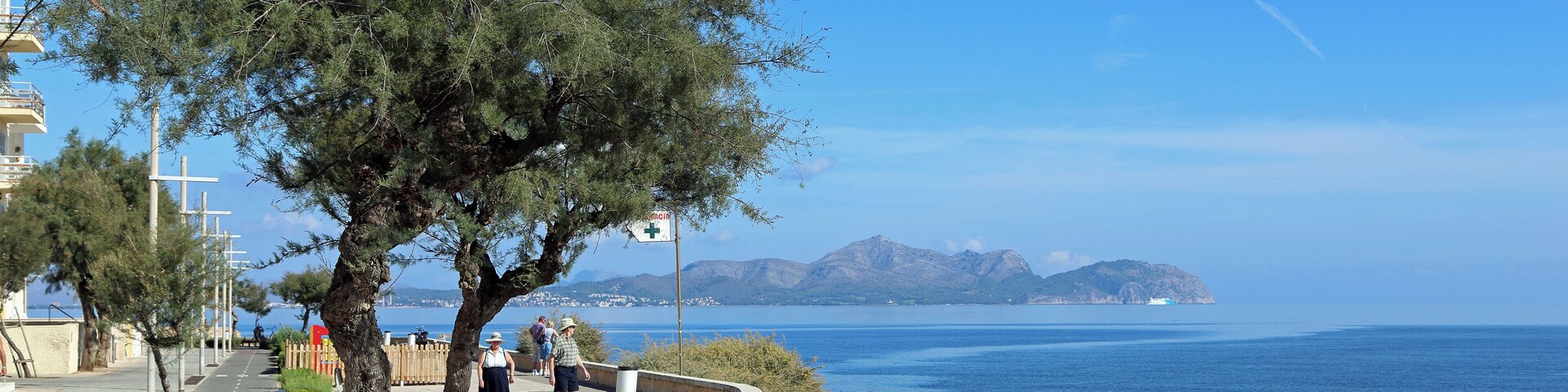 Uma passarela costeira com árvores, bancos e vista para o mar e montanhas ao longe.