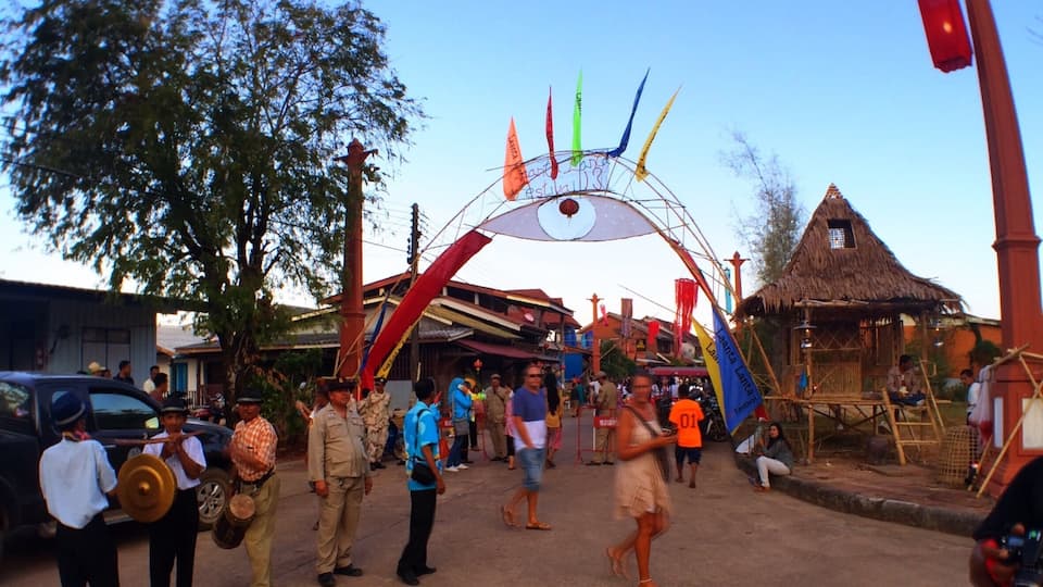 Cena de rua com um grande arco decorado com bandeiras coloridas e um desenho de olho. Há pessoas reunidas ao redor, algumas segurando bandeiras, e uma cabana com telhado de palha ao fundo.