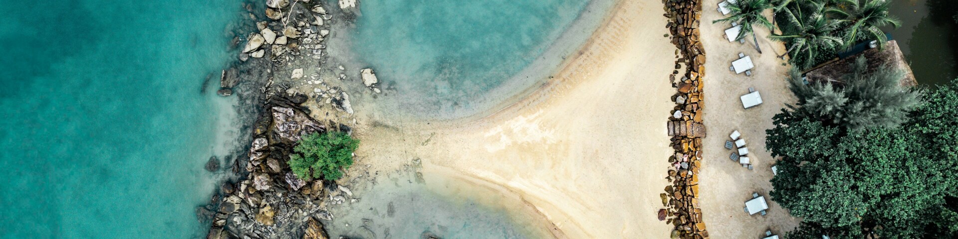 Uma praia com águas turquesa cristalinas, um afloramento rochoso e uma fileira de cadeiras de praia brancas.