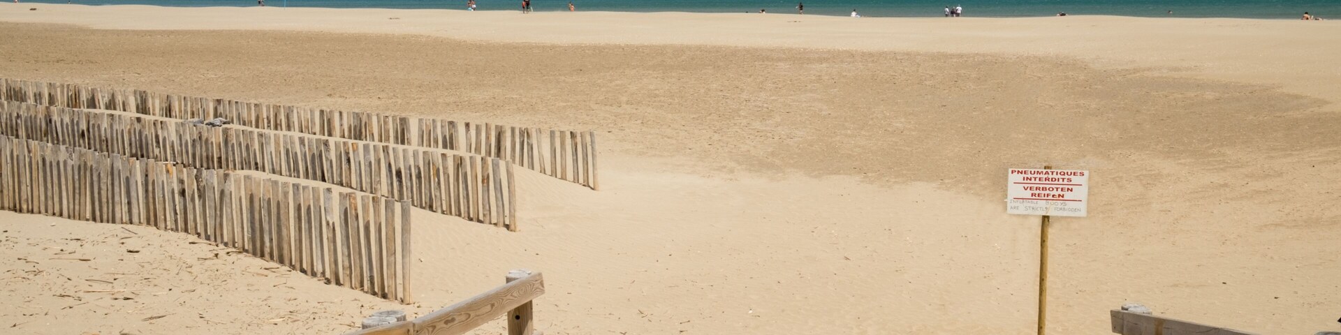 Um calçadão de madeira que leva a uma praia de areia, com céu azul claro e mar calmo.