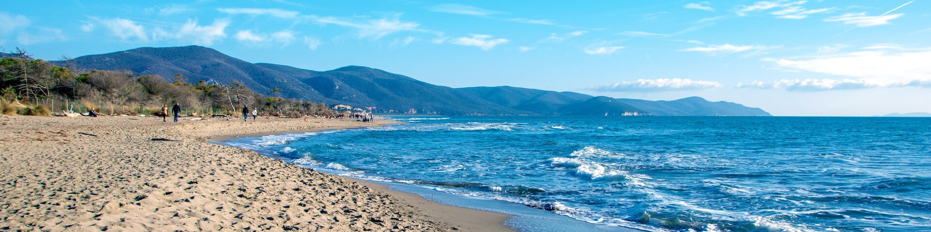 Uma praia arenosa com ondas, céu azul límpido e montanhas ao longe.
