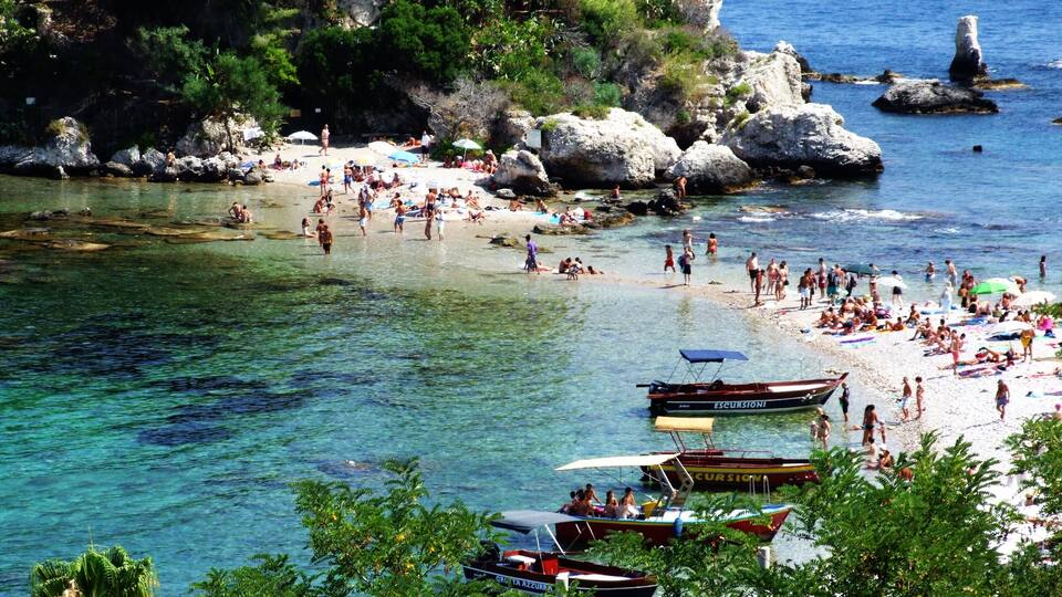 Uma praia movimentada, com pessoas se banhando e tomando sol, rodeada por falésias rochosas e águas azul-claras.