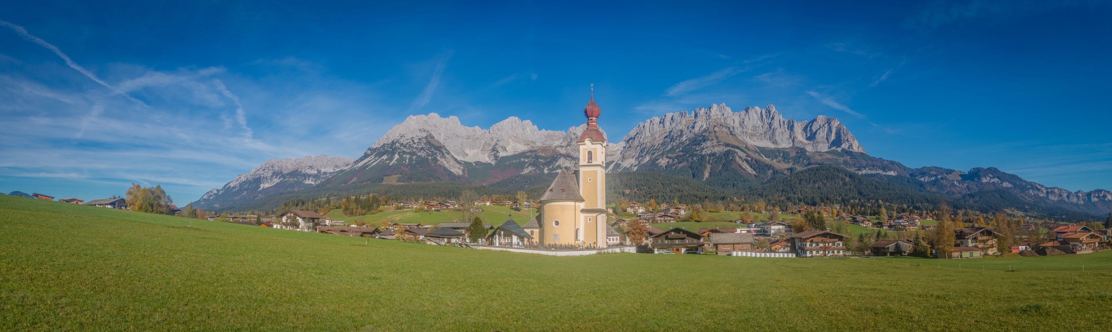 Uma igreja com cúpula vermelha e torre do sino, rodeada por uma aldeia e montanhas.