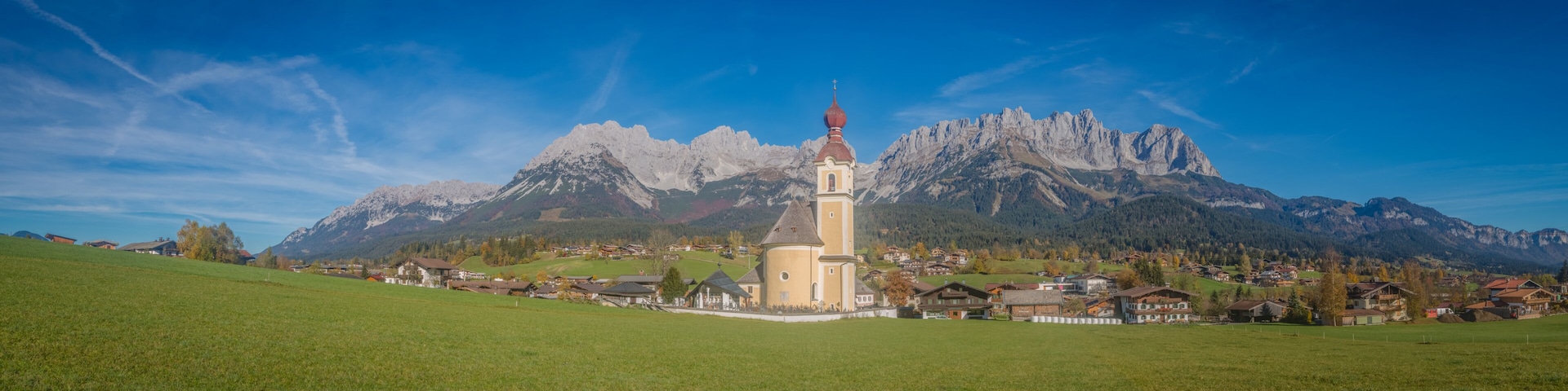 Uma igreja com cúpula vermelha e torre do sino, rodeada por uma aldeia e montanhas.