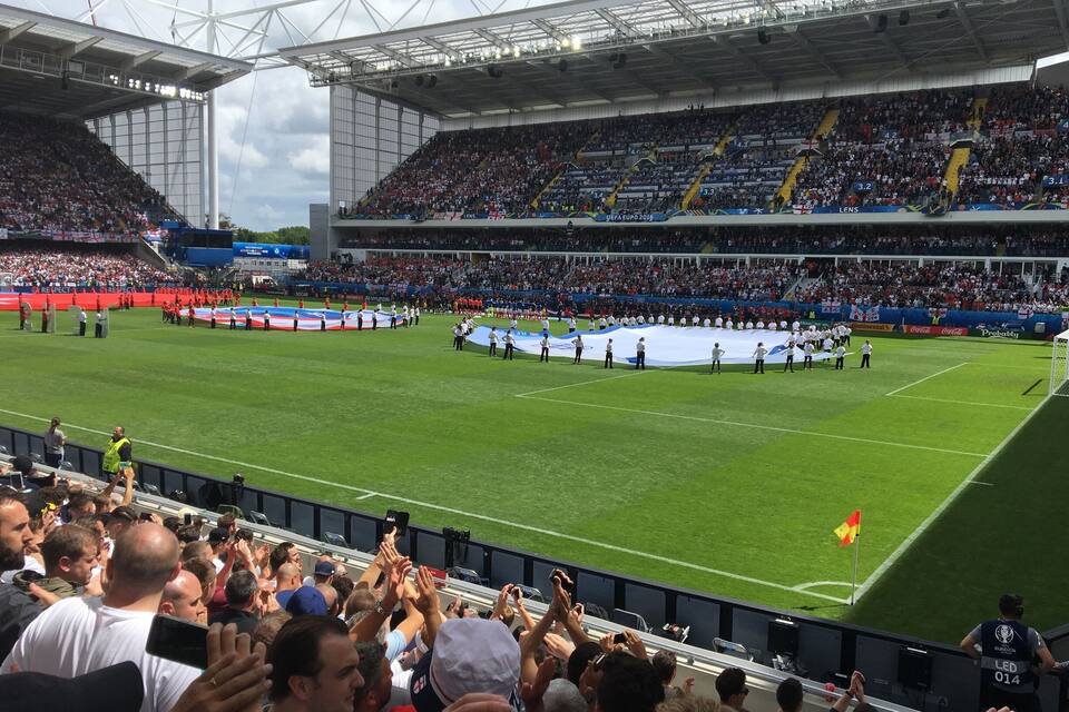 Um estádio lotado de espectadores, uma grande bandeira sendo hasteada e um placar parcialmente visível.