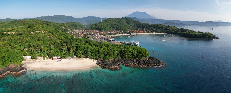 Uma cidade costeira com uma praia, águas turquesa cristalinas e colinas verdes exuberantes ao fundo.