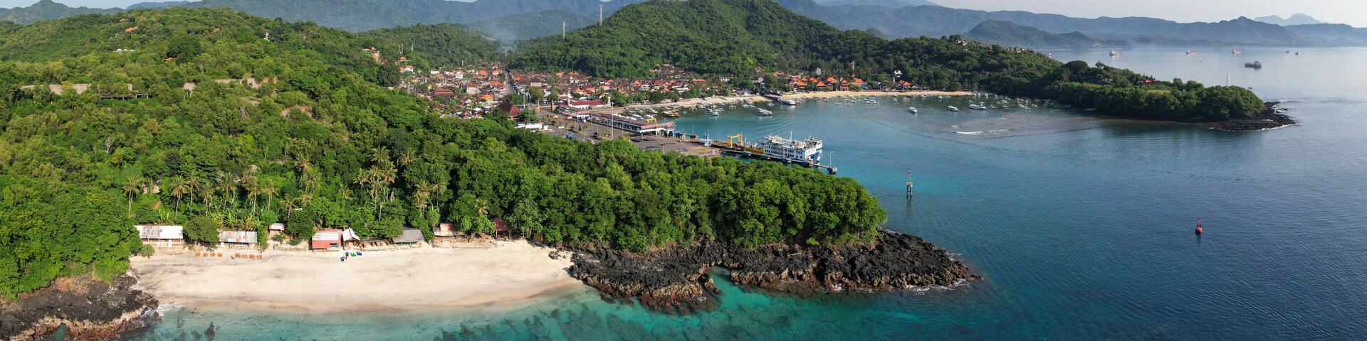 Uma cidade costeira com uma praia, águas turquesa cristalinas e colinas verdes exuberantes ao fundo.
