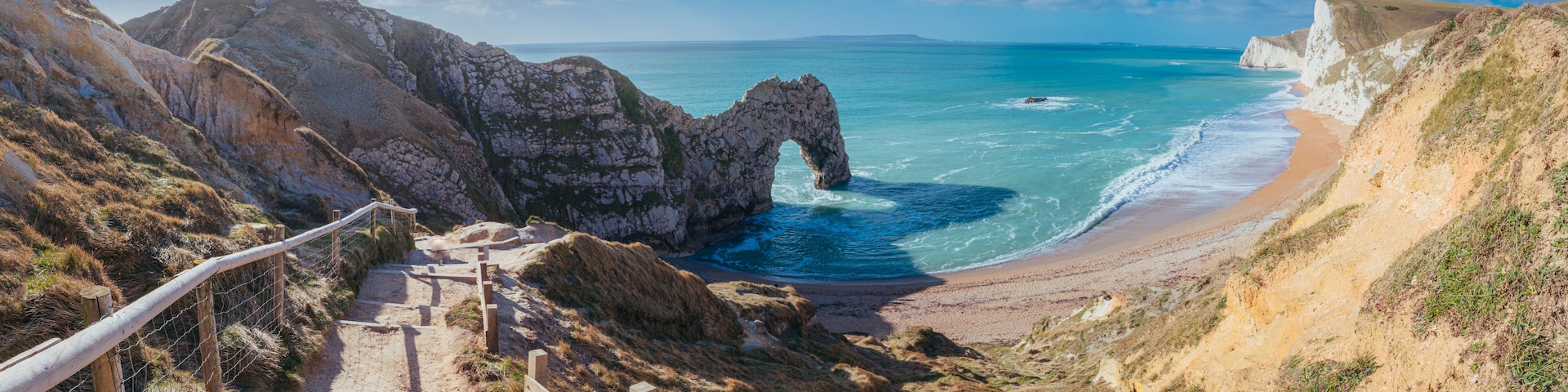 Paisagem costeira com um arco rochoso imponente, uma praia arenosa e um céu azul claro.