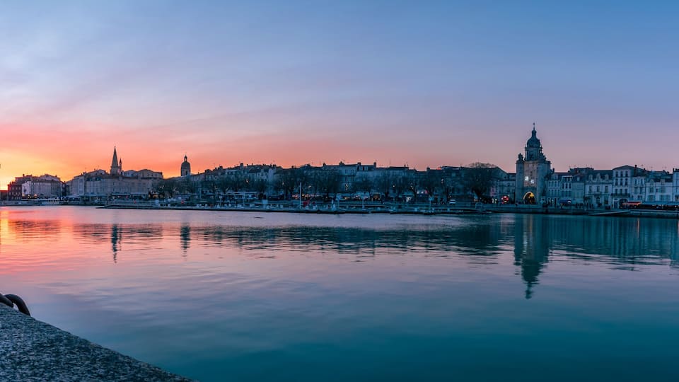 Um horizonte urbano ao pôr do sol, com um rio calmo refletindo os edifícios, uma torre imponente e uma ponte.