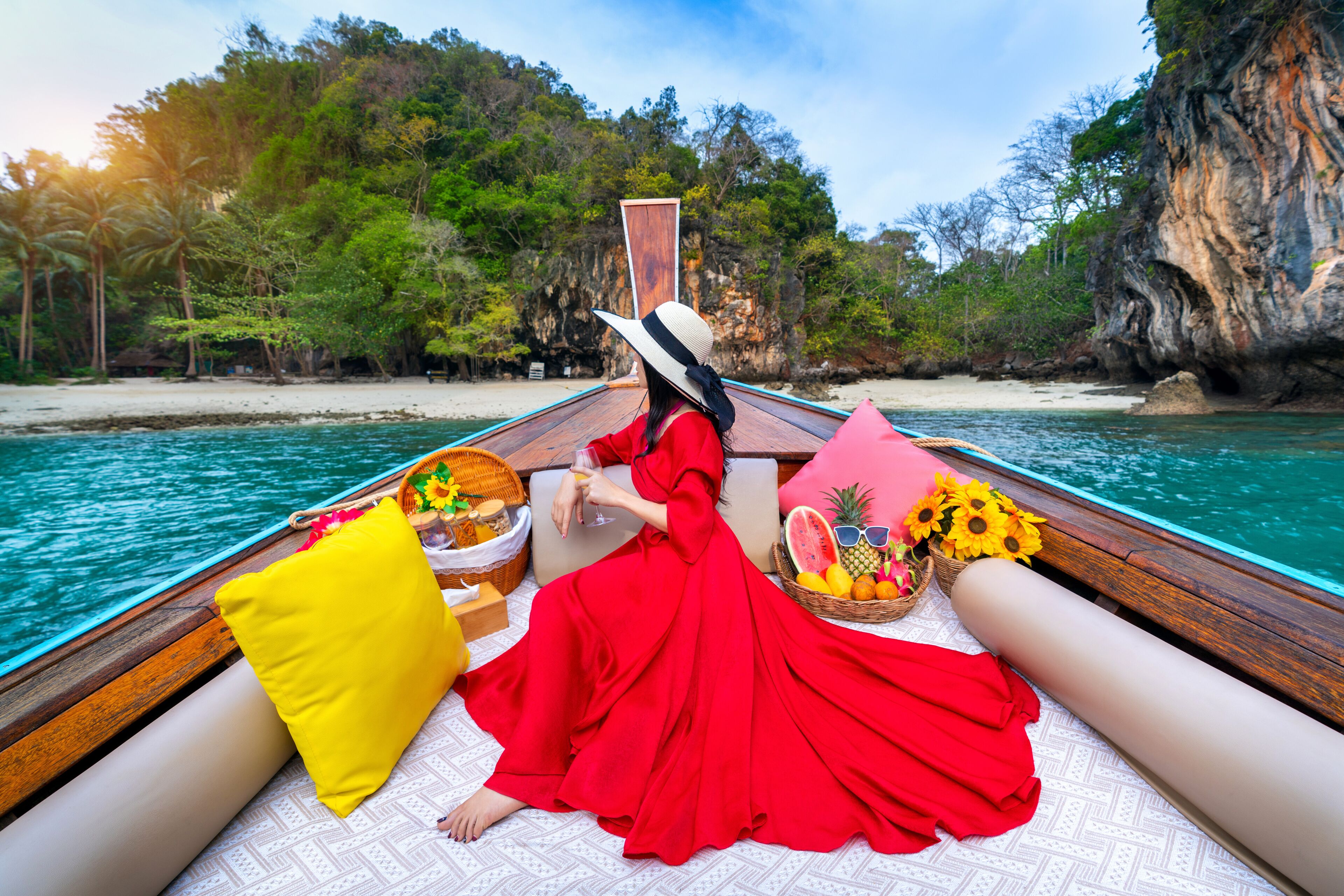 Mulher de vestido e chapéu vermelho em barco, com uma praia tropical e falésias ao fundo.