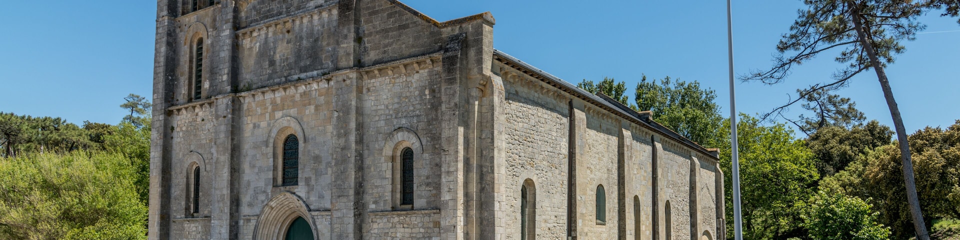 Uma igreja de pedra histórica com janelas em arco e um campanário, cercada por um pátio pavimentado e vegetação.
