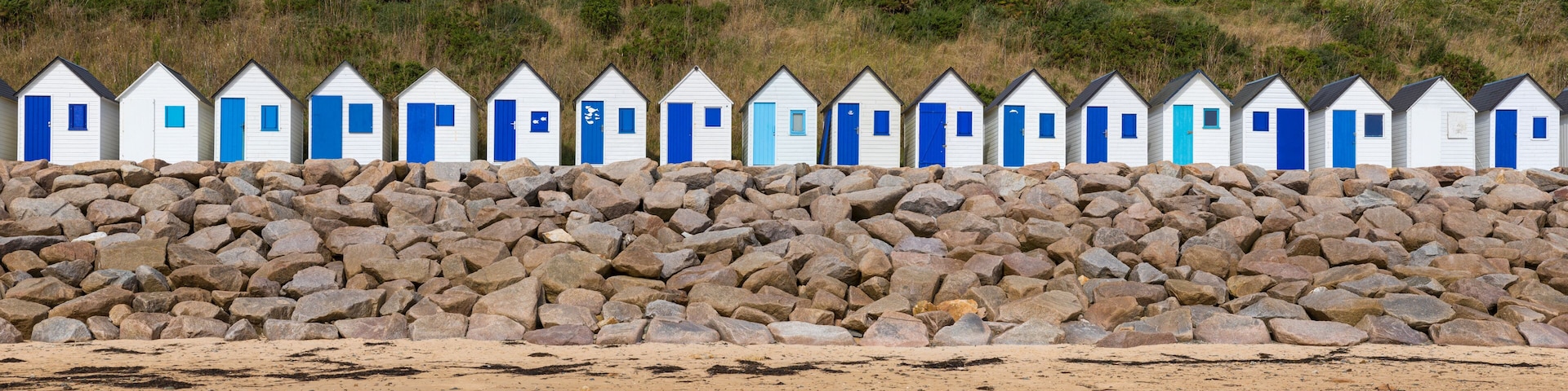 Uma fileira de cabanas de praia brancas, com portas e janelas azuis, alinhadas em frente a uma praia rochosa e uma colina gramada ao fundo.