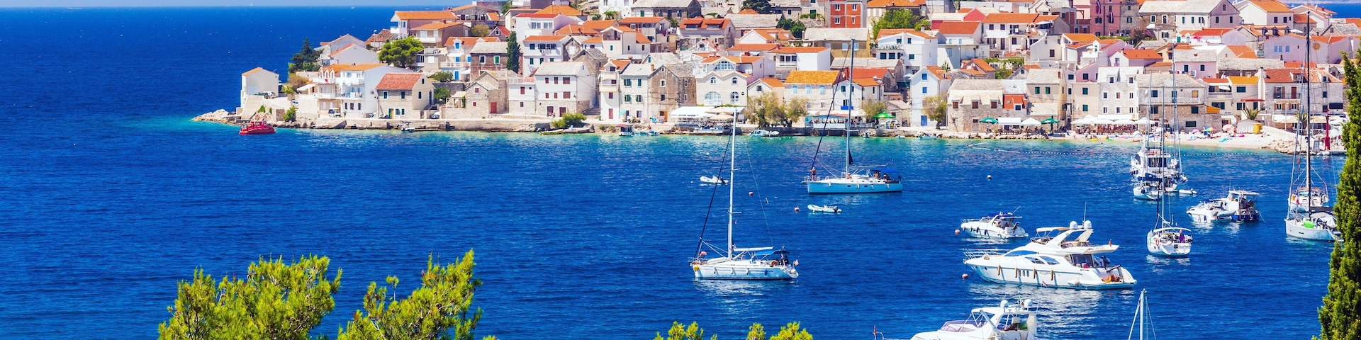 Uma vila costeira com uma torre imponente no topo de uma colina, mar azul cristalino e vários barcos ancorados no porto.