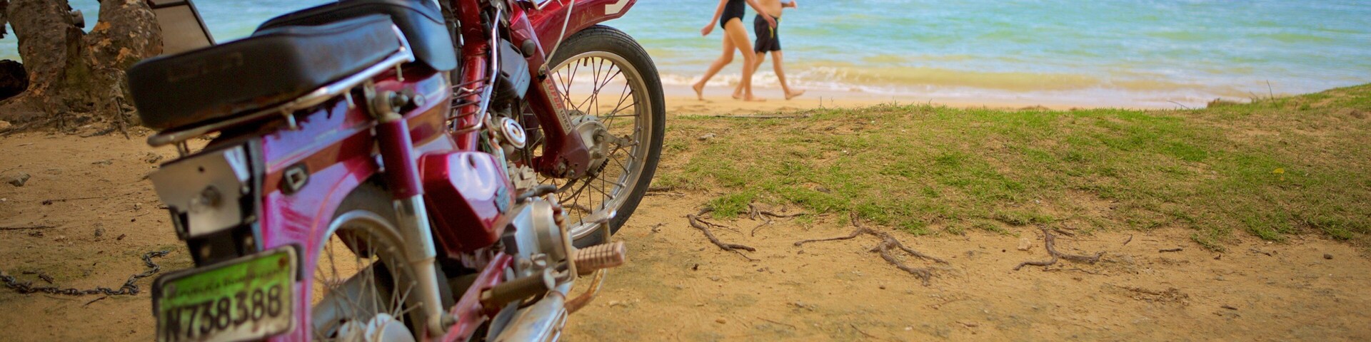 Uma motocicleta estacionada numa praia arenosa, com vista para o oceano e um veleiro ao longe.