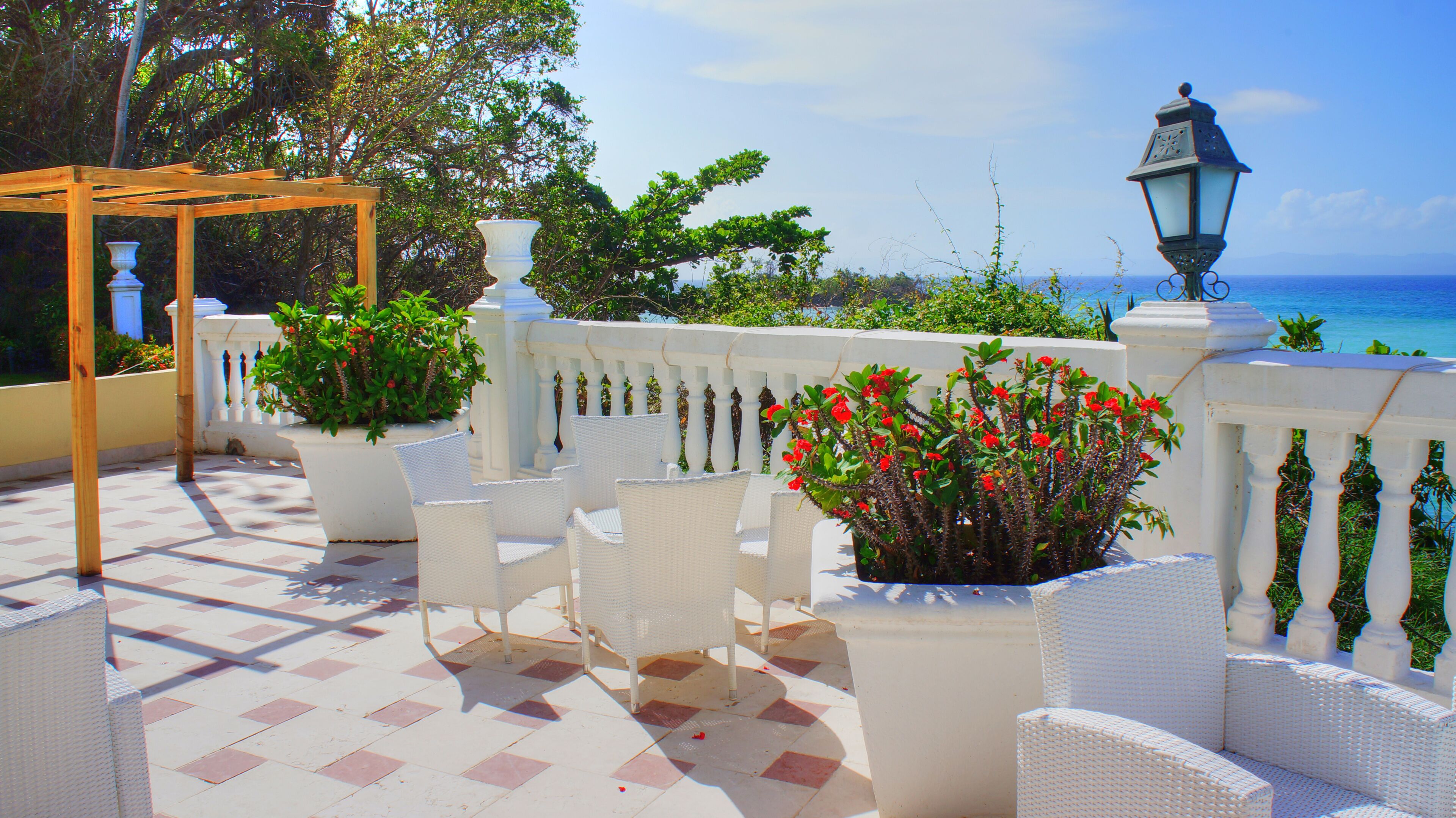 Um pátio à beira-mar com mobiliário de vime branco, plantas em vasos e um candeeiro decorativo.