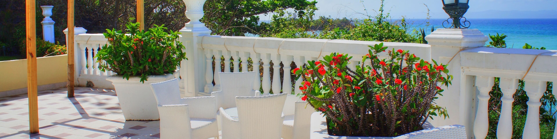 Um pátio à beira-mar com mobiliário de vime branco, plantas em vasos e um candeeiro decorativo.