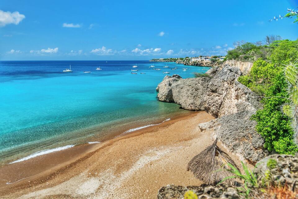 Uma praia com águas turquesa cristalinas, costa rochosa e barcos ancorados.