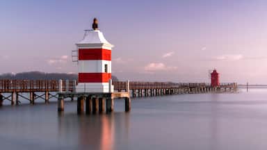Um farol vermelho e branco numa doca que se estende por águas calmas, com um segundo farol ao longe e uma linha de costa arborizada.