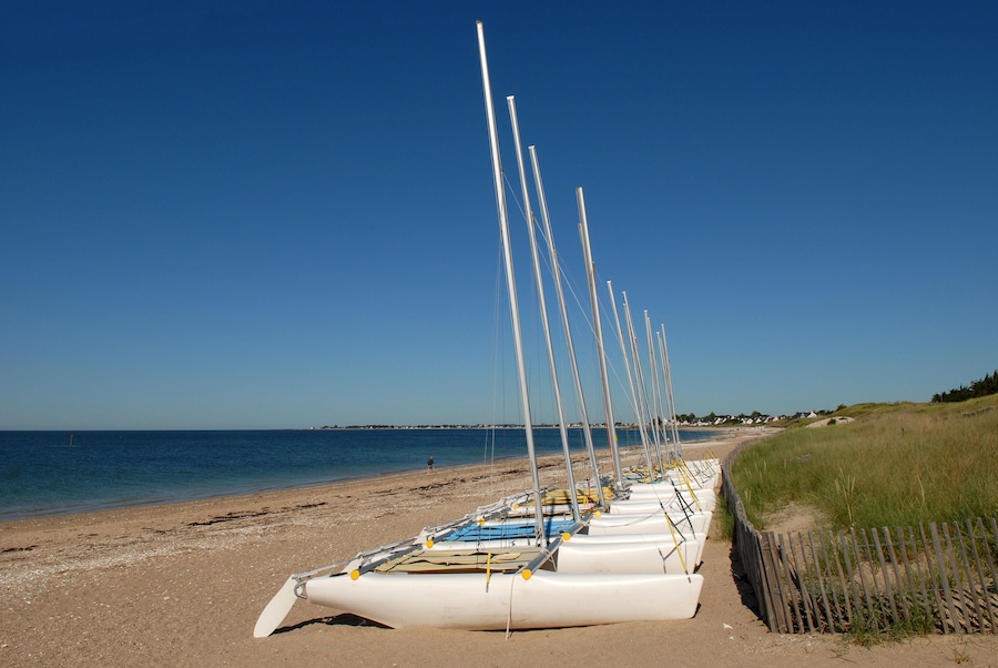 Uma fileira de barcos à vela alinhados em uma praia arenosa, com o oceano ao fundo.