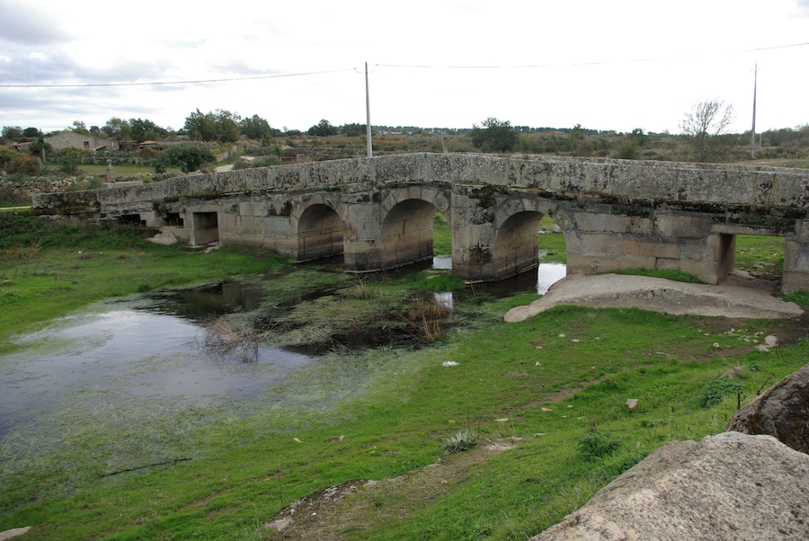 Uma ponte de pedra antiga sobre um rio pequeno, com margens gramadas e uma aldeia ao longe.