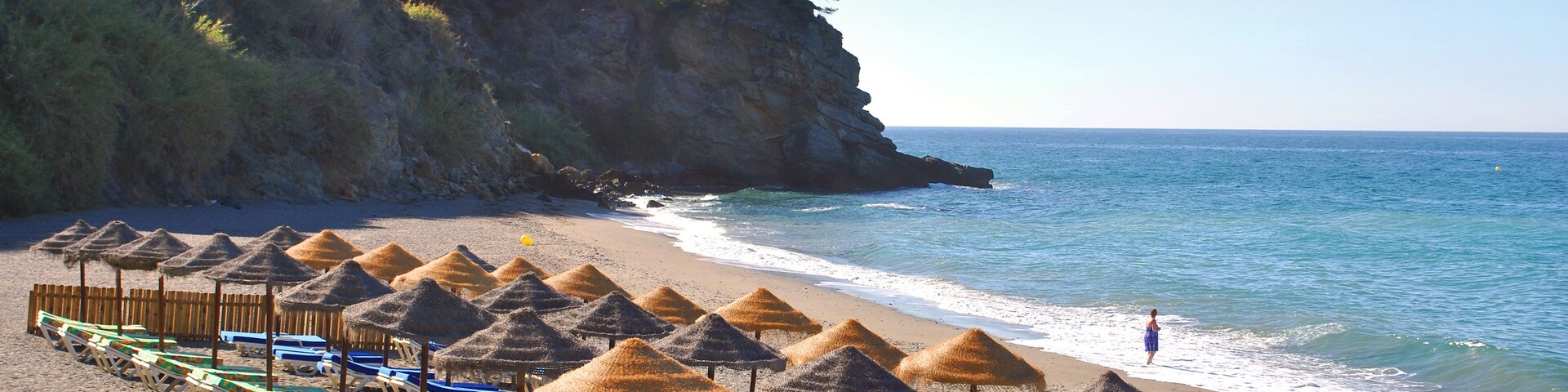 Uma praia com fileiras de guarda-sóis e espreguiçadeiras, um penhasco rochoso e o oceano.