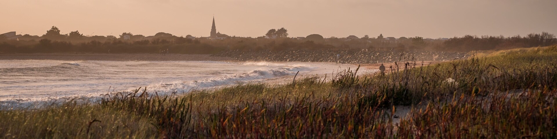 Paisagem costeira com ondas, dunas gramadas e o topo de uma igreja ao longe.