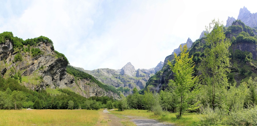 Paisagem montanhosa com trilha de terra, floresta verde densa e penhascos rochosos.