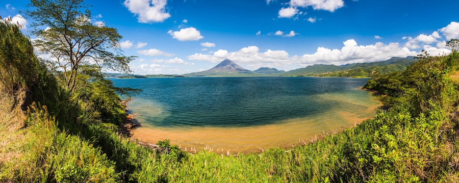 Paisagem costeira com vista clara para uma montanha ao longe, rodeada por vegetação exuberante e um mar calmo.