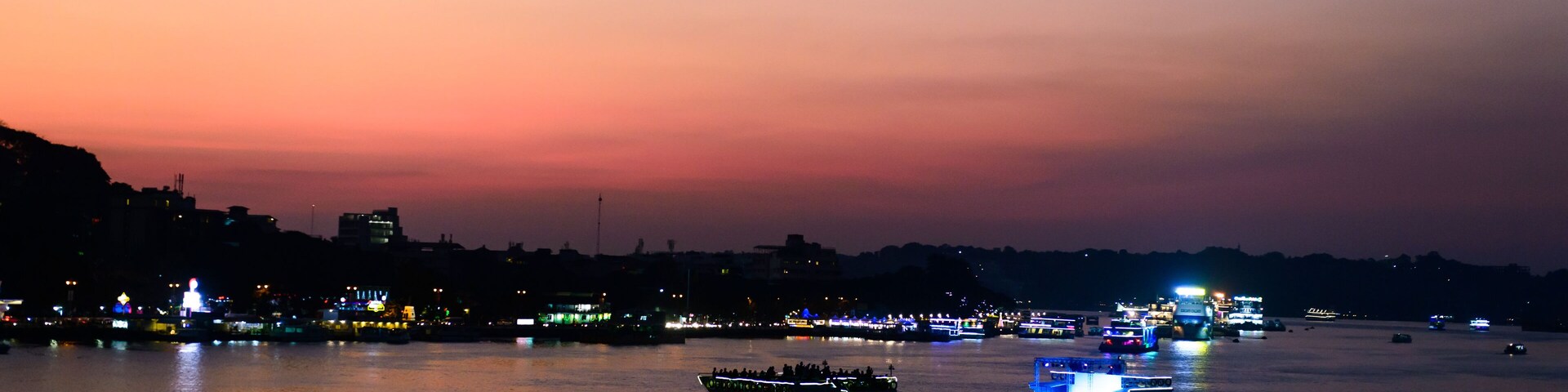 Um rio ao pôr do sol, com barcos iluminados e o horizonte da cidade ao fundo.