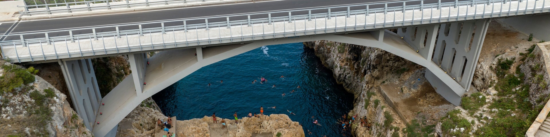 Uma ponte sobre um litoral rochoso, com pessoas nadando nas águas abaixo.