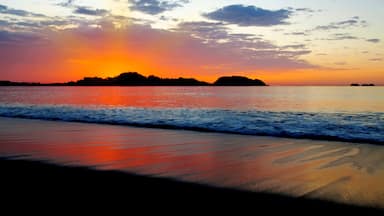 Um pôr do sol sobre uma praia com ondas, uma ilha ao longe e céu parcialmente nublado.