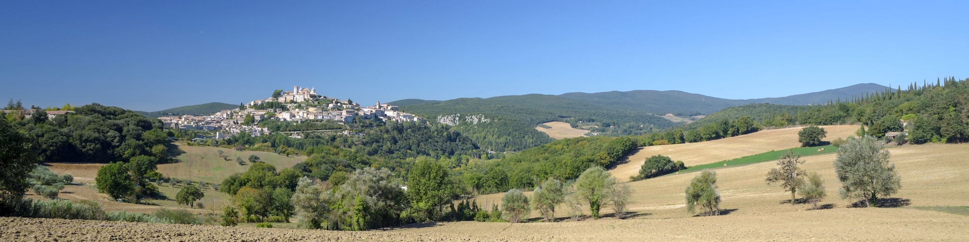 Uma aldeia no alto de uma colina, com edifícios, rodeada por colinas e campos verdes.