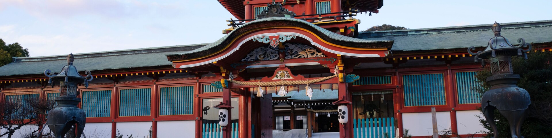 Um templo japonês tradicional com telhado vermelho e verde, uma grande estátua de uma divindade e uma escadaria de pedra que leva à entrada.