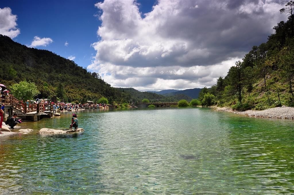 Um rio límpido cercado por colinas arborizadas, uma ponte de madeira e pessoas aproveitando a água.