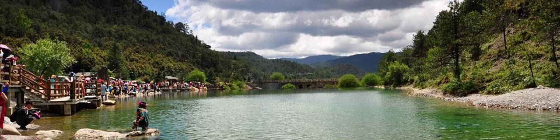 Um rio límpido cercado por colinas arborizadas, uma ponte de madeira e pessoas aproveitando a água.