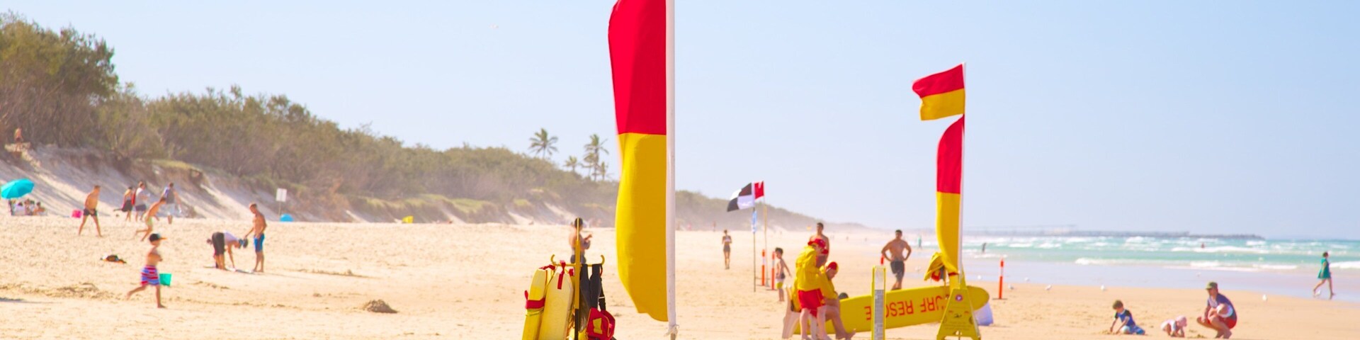 Bandeiras de salva-vidas, um prancha de surf amarela e pessoas em uma praia arenosa.