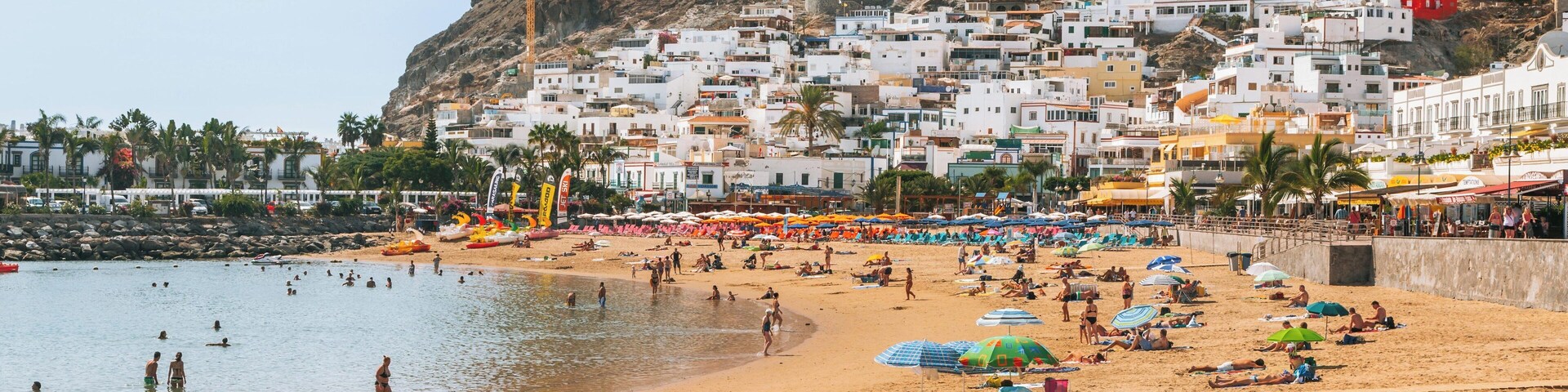 Uma praia lotada com guarda-sóis coloridos e pessoas tomando sol. Ao fundo, um morro rochoso com edifícios brancos.
