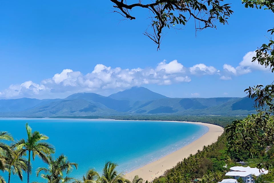 Paisagem costeira com mar azul claro, uma praia de areia e colinas verdejantes ao fundo.