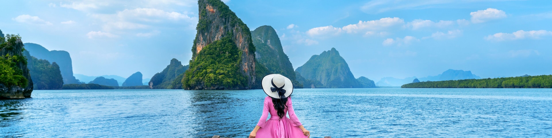 Uma mulher de vestido rosa e chapéu de aba larga está em um píer de madeira, olhando para um corpo de água com ilhas verdes e uma grande formação rochosa ao longe.